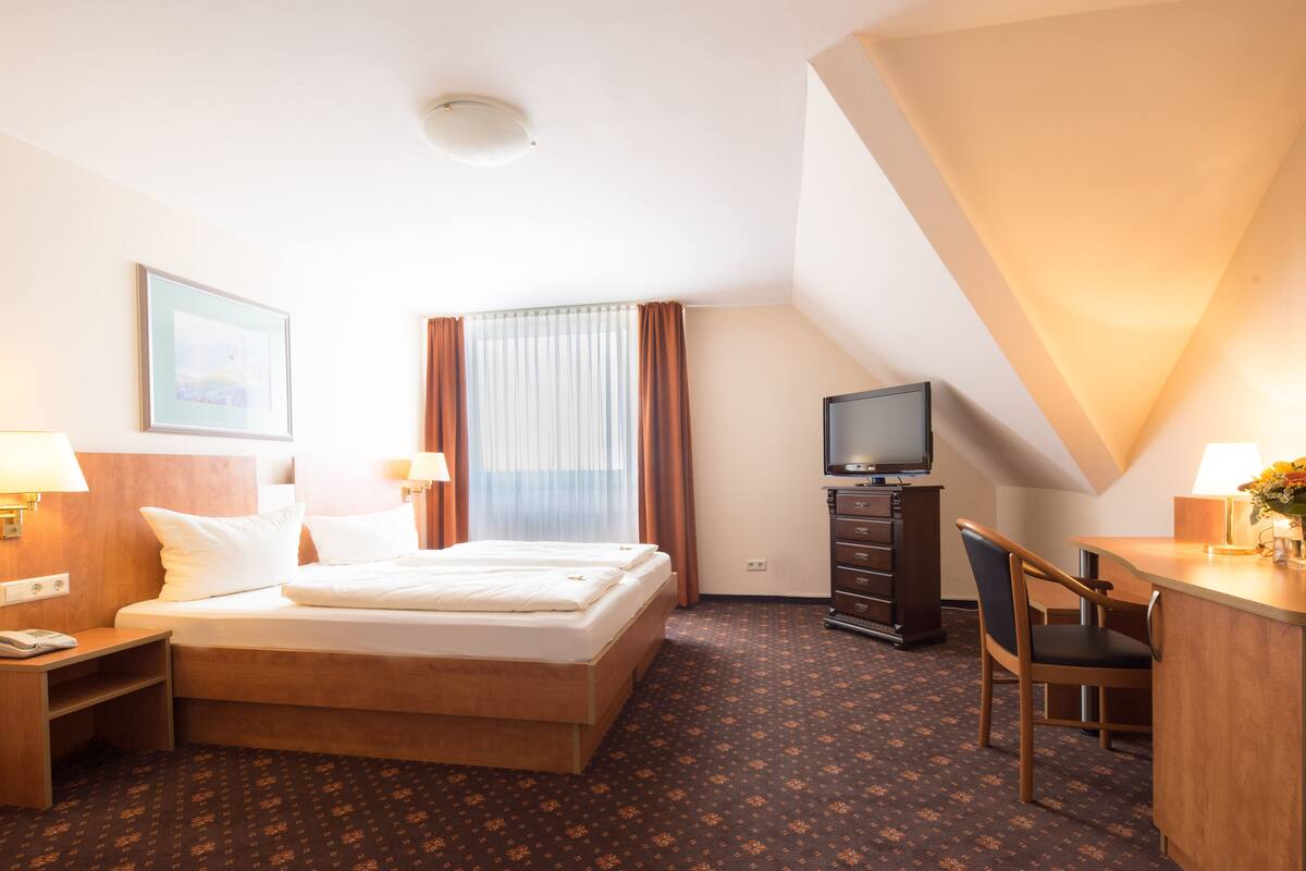 Quarto de hotel com cama, escrivaninha, cadeira, televisão e janela com cortinas.