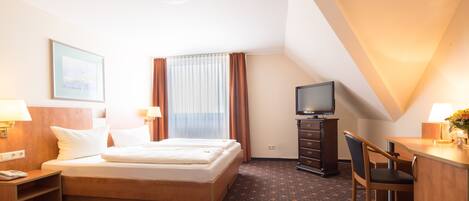 Quarto de hotel com cama, escrivaninha, cadeira, televisão e janela com cortinas.