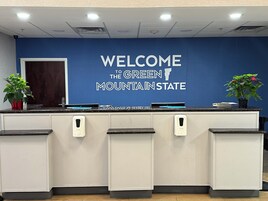 Bancada de recepção com parede azul exibindo a placa "WELCOME TO THE GREEN MOUNTAIN STATE". Há duas plantas em vasos no balcão.