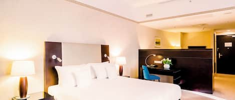 Quarto de hotel com uma cama grande, uma escrivaninha com cadeira, uma luminária e uma pequena planta.
