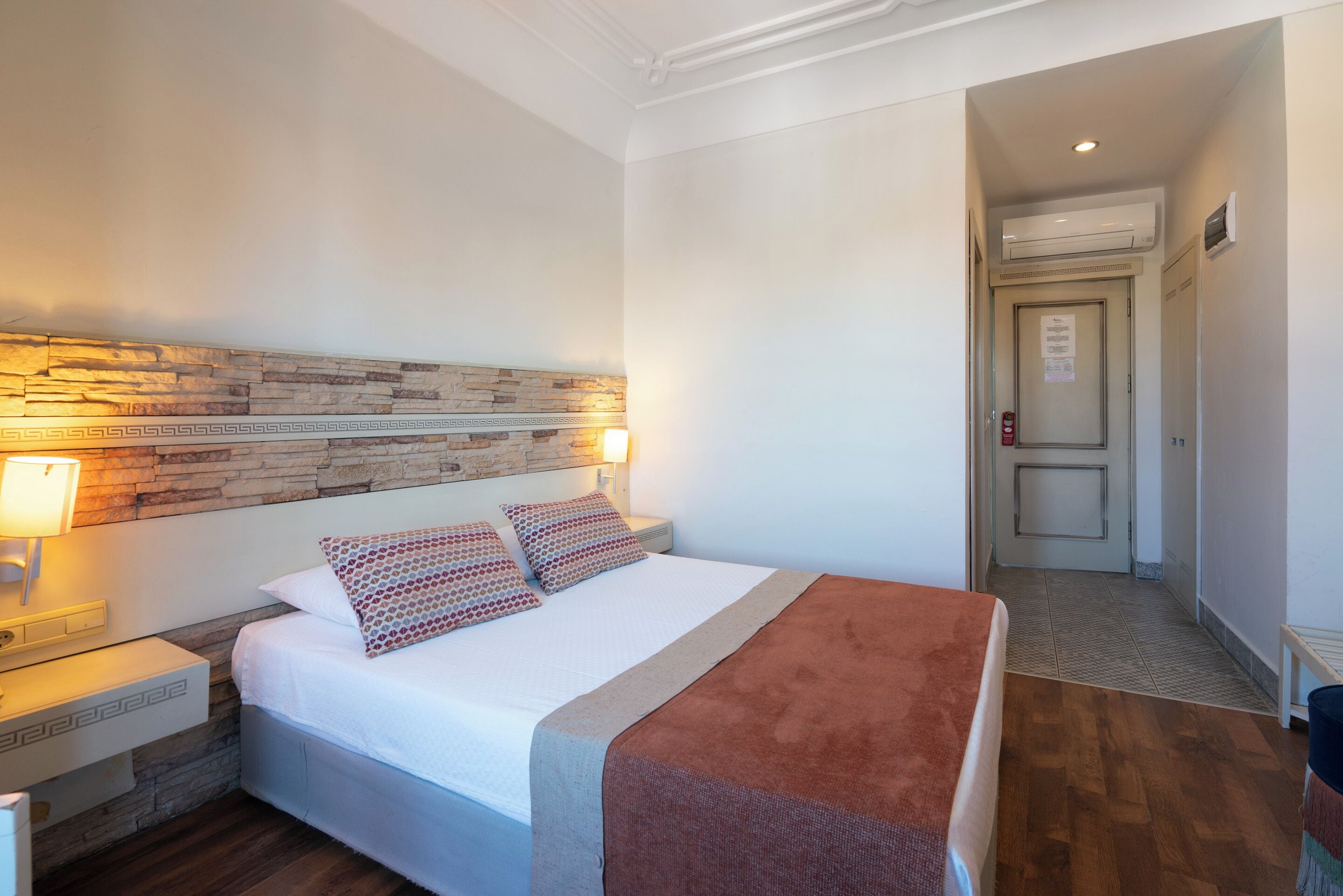Quarto de hotel com cama, mesa de cabeceira, luminária na parede e cabeceira de madeira.