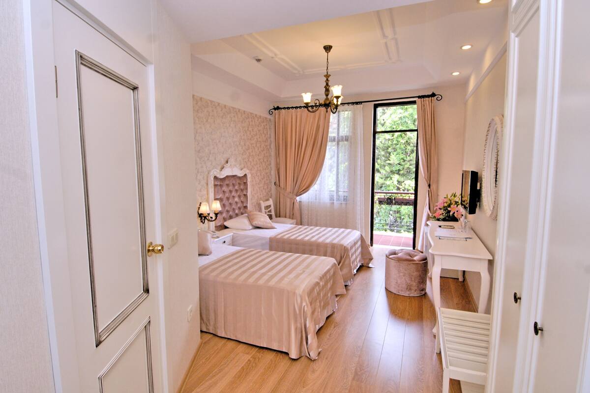 Quarto com duas camas, uma escrivaninha, uma cadeira, uma mesinha, um espelho e uma janela com cortinas.