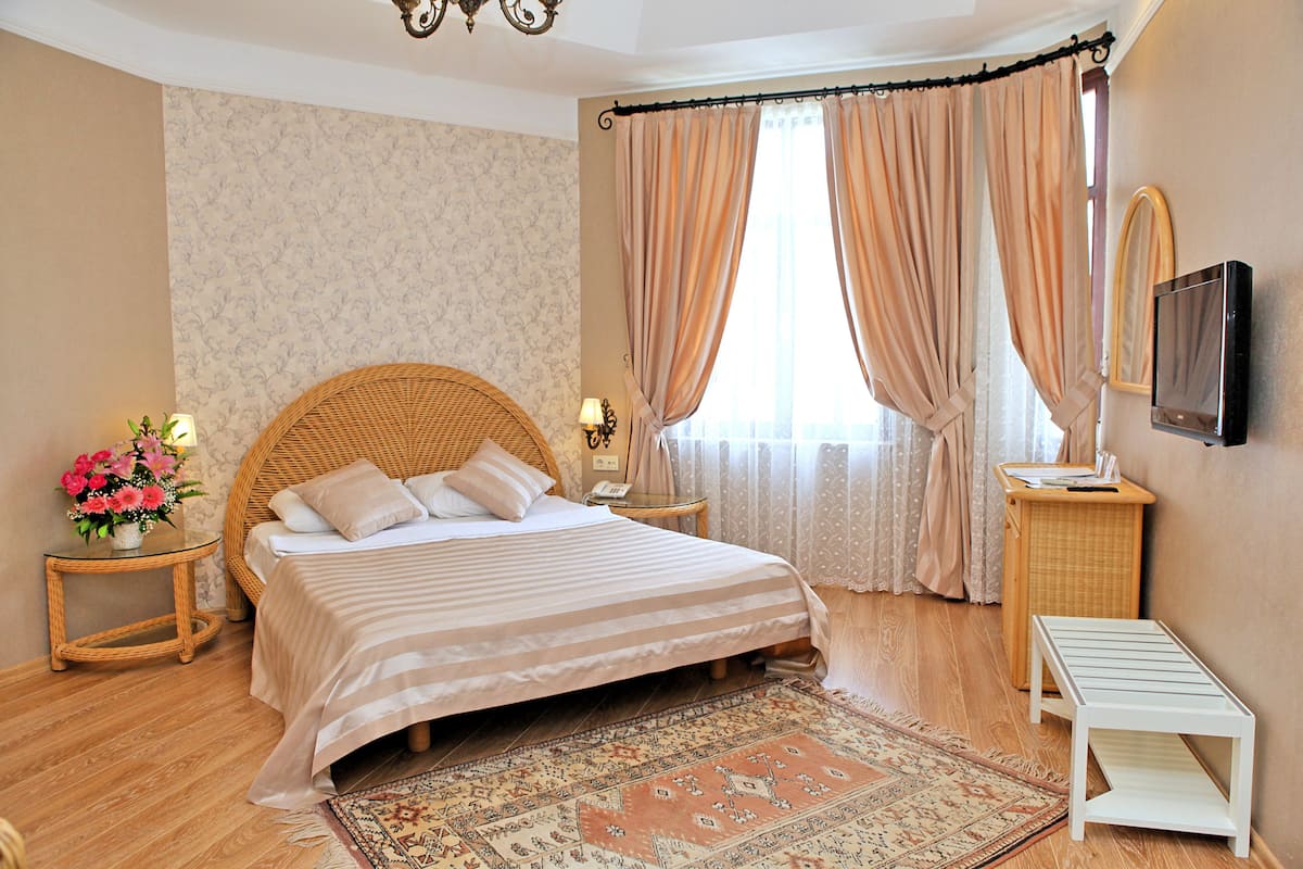 Quarto com uma cama grande, mesa de cabeceira com abajur, pequena mesa lateral com um vaso de flores, televisão na parede e janela com cortinas.