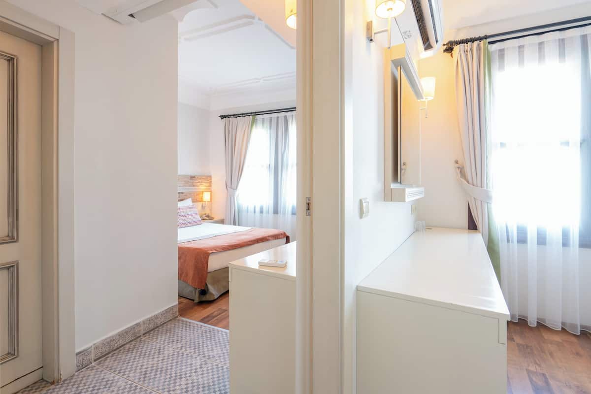 Quarto de hotel com uma cama, uma pequena escrivaninha, um espelho e uma janela com cortinas.