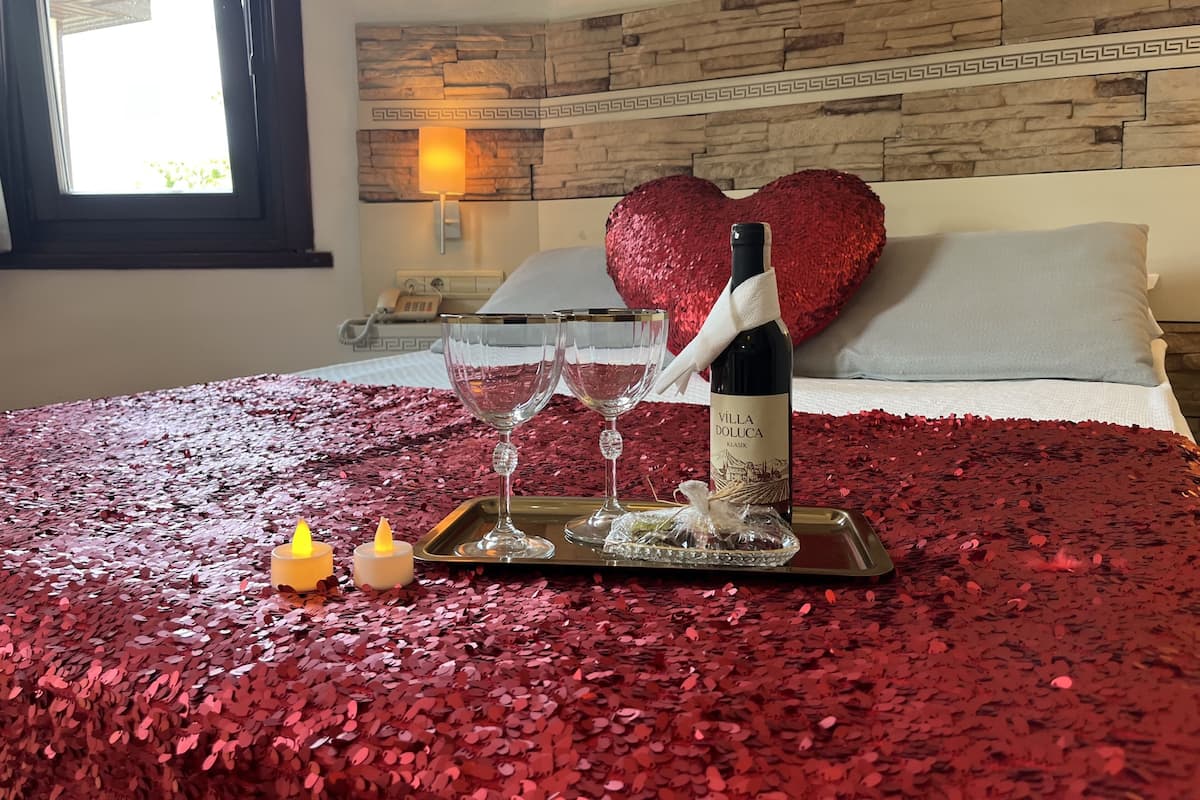 Quarto de hotel com uma cama, parede decorativa, travesseiro em formato de coração, uma garrafa de vinho, duas taças e um tabuleiro com velas.