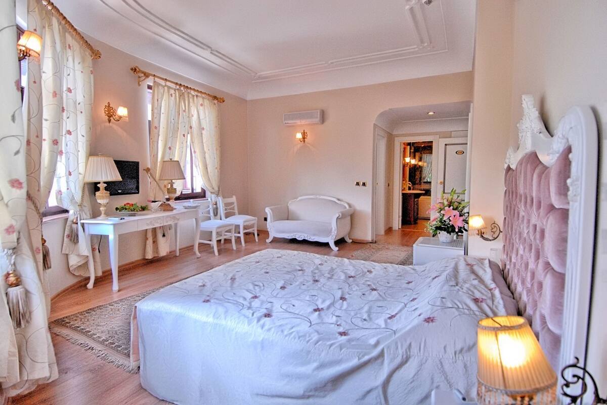 Quarto com cama, escrivaninha, cadeiras, penteadeira e banheiro visível por uma porta aberta.