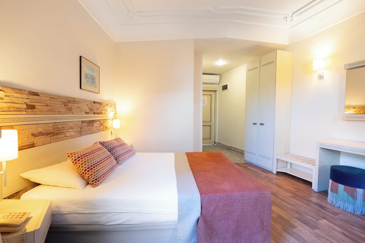 Quarto de hotel com cama, cabeceira de madeira, mesa de cabeceira, luminária, um banco e uma pequena mesa com luminária.