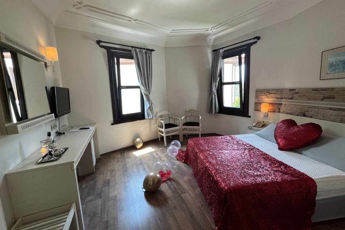 Quarto de hotel com cama, escrivaninha, cadeira, televisão e janela com cortinas.