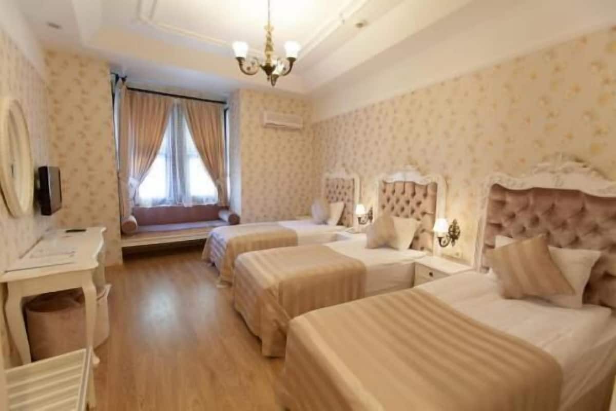 Quarto de hotel com duas camas, uma escrivaninha, uma cadeira, janela com cortinas e um lustre.