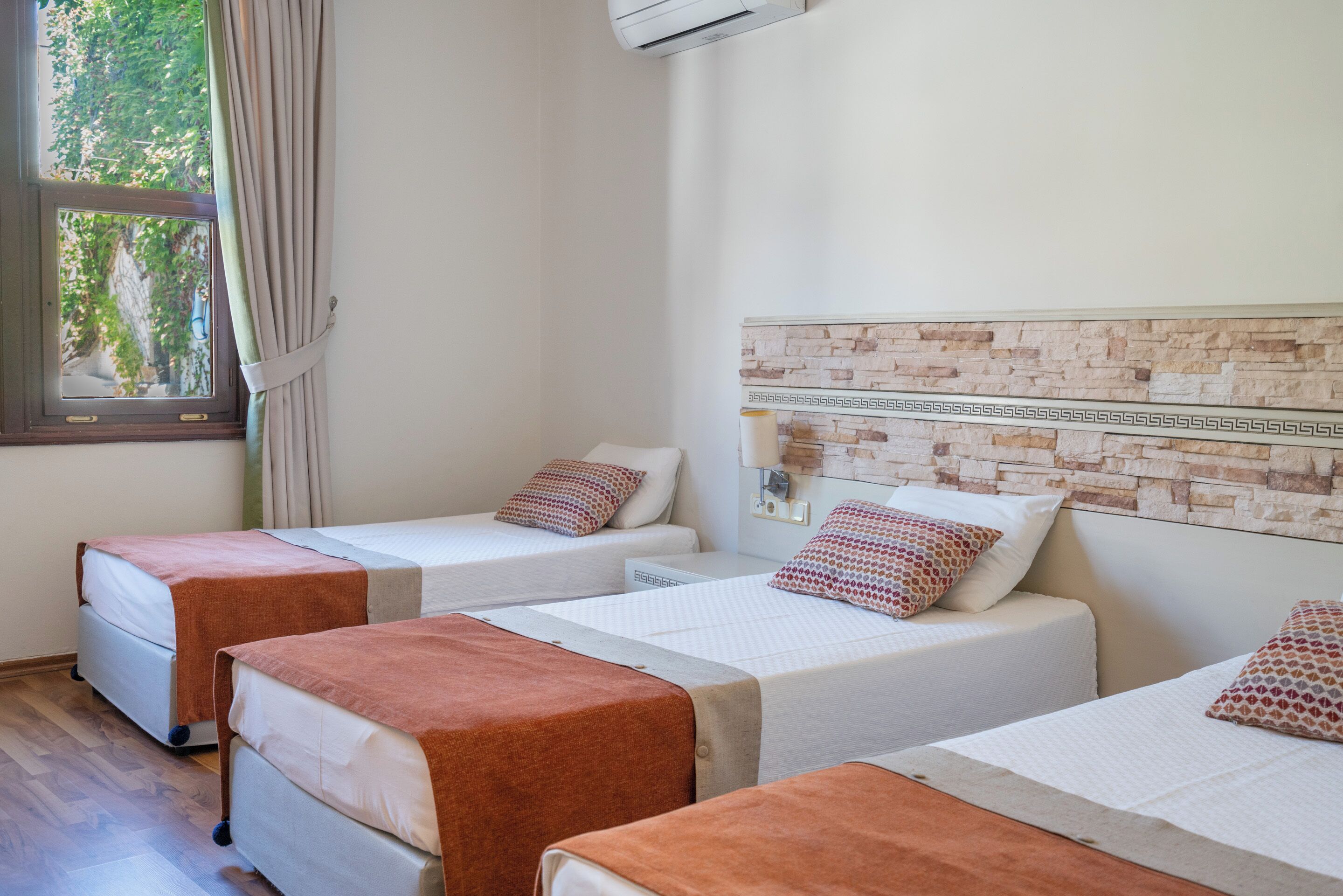 Quarto de hotel com três camas de solteiro, cada uma com um cobertor marrom e almofadas estampadas. Há uma janela com cortinas, um aparelho de ar condicionado acima dela e uma parede com padrão de pedra.