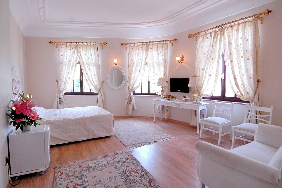 Quarto de hotel com cama, escrivaninha, duas cadeiras, televisão e mesinha de cabeceira com abajur.