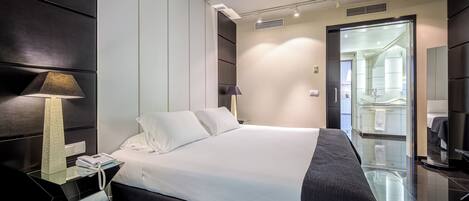 Quarto de hotel moderno com uma cama grande, mesas de cabeceira, uma luminária e um banheiro visível por uma porta aberta.