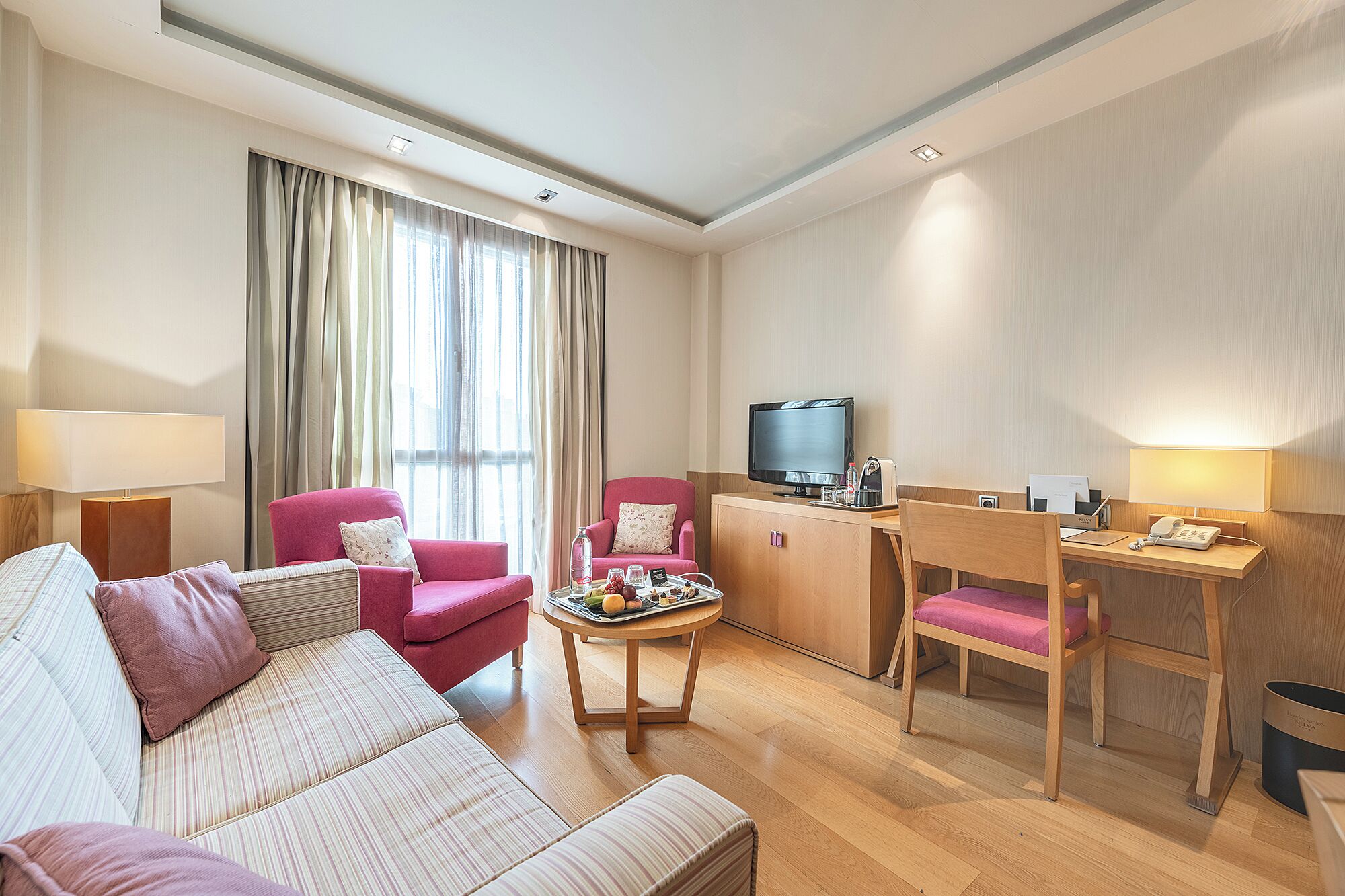 Quarto de hotel com sofá, duas poltronas, mesa de jantar com cadeiras, televisão e escrivaninha com abajur.