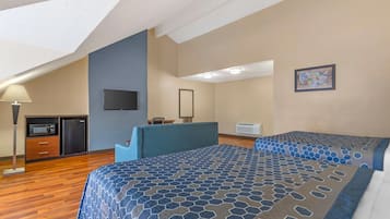 Quarto de hotel com teto alto, cama com colcha estampada, escrivaninha com cadeira, televisão de tela plana na parede e pequena área de kitchenette.