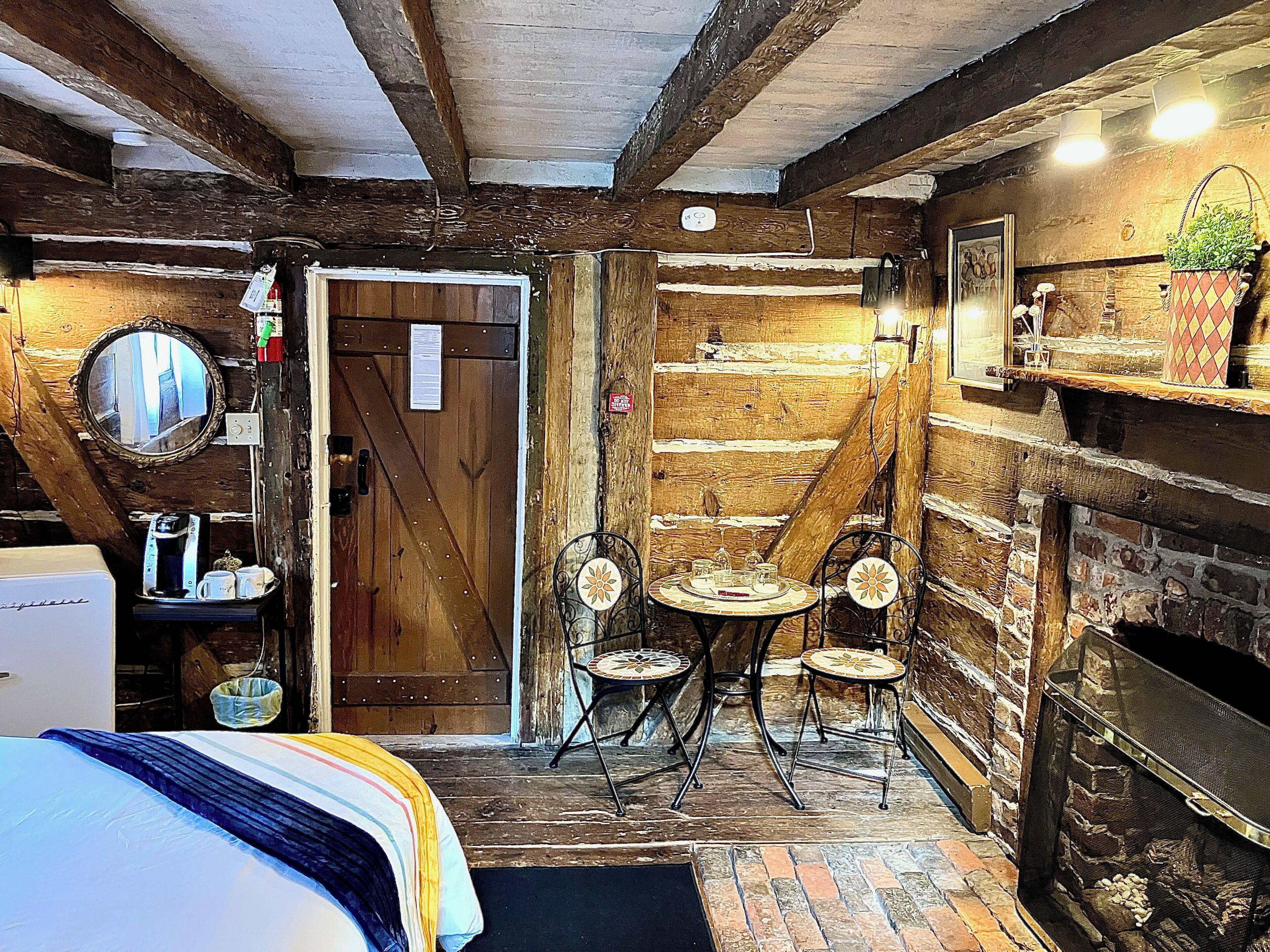 Quarto rústico com uma cama, porta de madeira, pequena mesa com cadeiras, espelho e lareira.