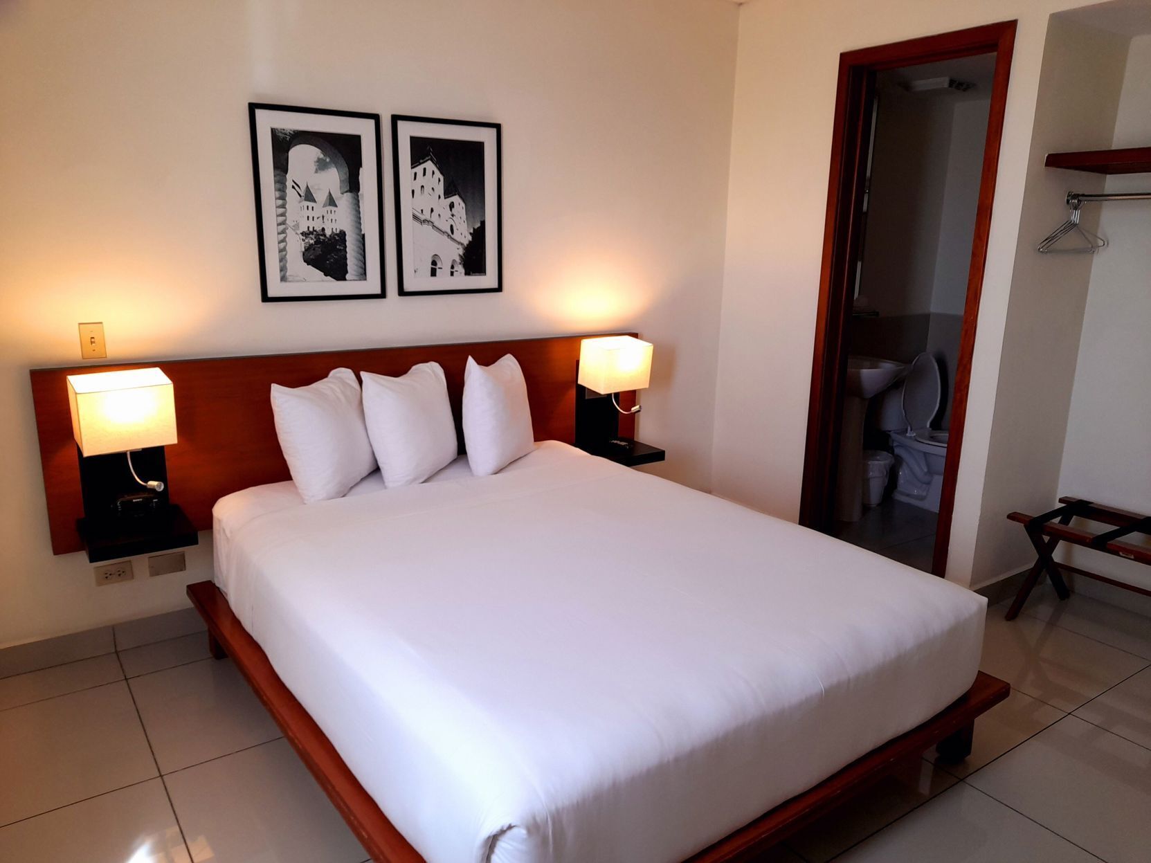 Quarto de hotel com uma cama grande, duas luminárias de cabeceira, um painel de cama de madeira e duas quadros na parede.