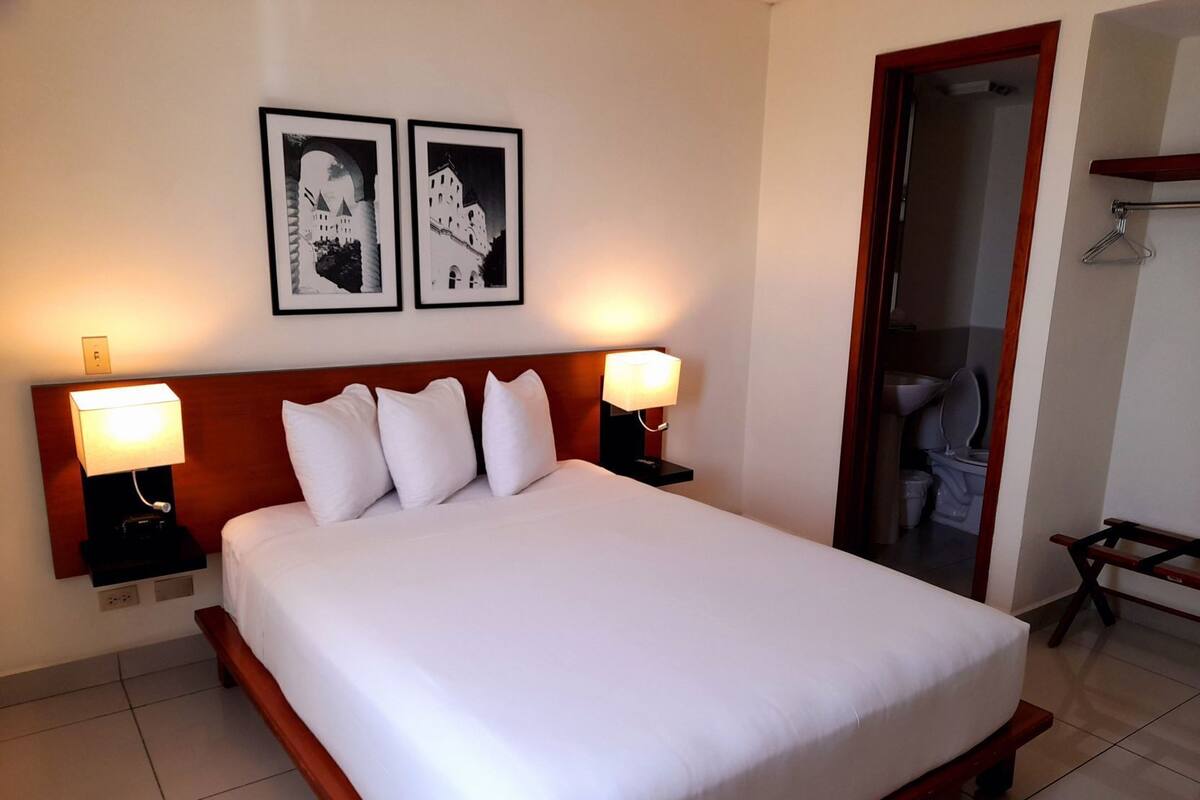 Quarto de hotel com uma cama grande, duas luminárias de cabeceira, um painel de cama de madeira e duas quadros na parede.