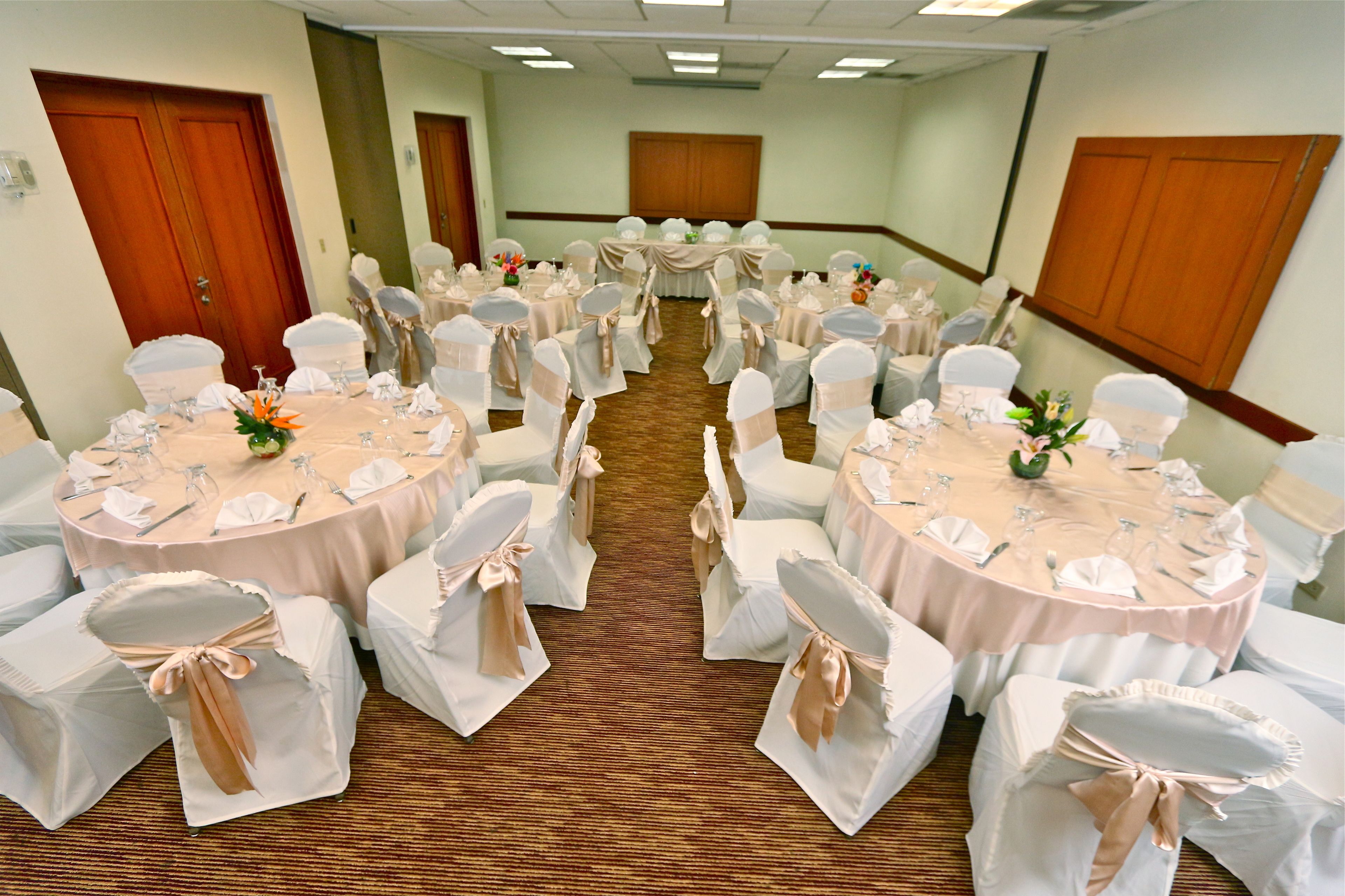 Uma sala de banquetes com mesas redondas dispostas para refeições, cada uma com cadeiras brancas e centros de mesa.