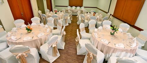 Uma sala de banquetes com mesas redondas dispostas para refeições, cada uma com cadeiras brancas e centros de mesa.