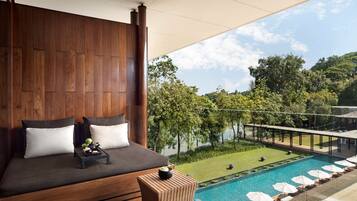Quarto moderno com cabeceira de madeira, um sofá com almofadas, uma mesinha com frutas e vista para uma piscina e jardim.