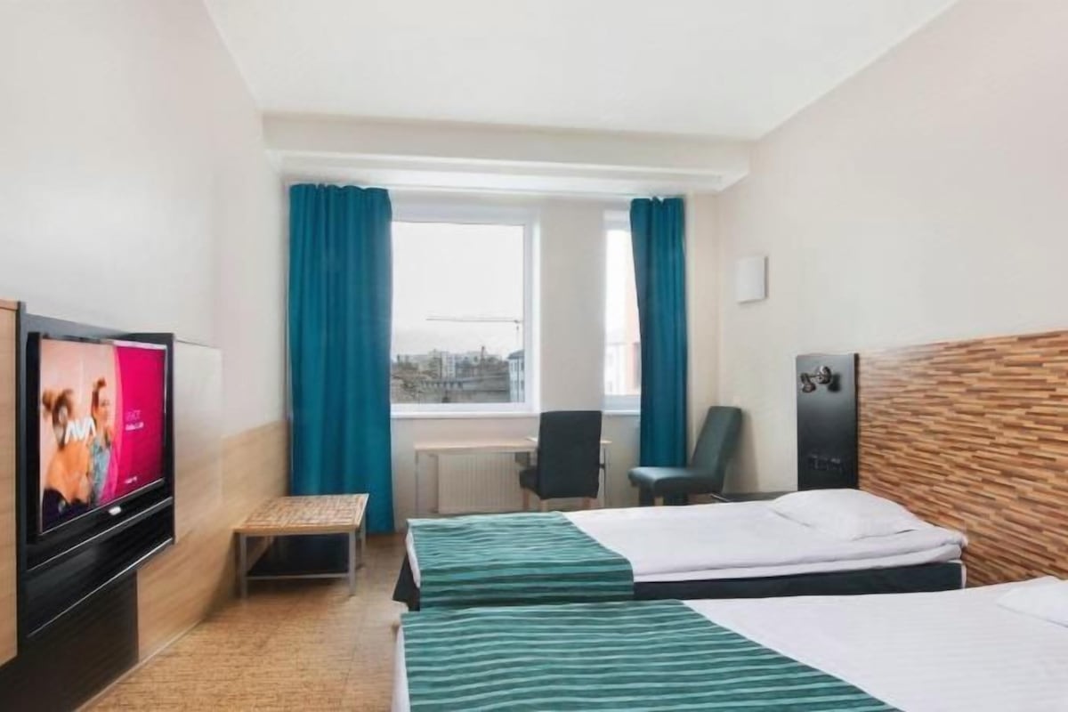 Quarto de hotel com cama, escrivaninha, cadeira, televisão e janela com cortinas.