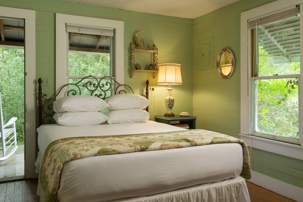 Quarto com cama, mesa de cabeceira, abajur, espelho e janela.