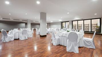 Um amplo salão de banquetes com várias mesas redondas e cadeiras brancas, janelas grandes e um piso de madeira envernizada.