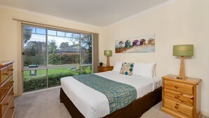 Um quarto com uma janela grande, uma cama com colcha estampada, uma mesinha de cabeceira de madeira com abajur e um quadro na parede.