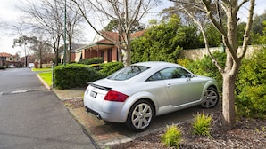 Um carro prateado estacionado numa rua residencial, com casas e árvores ao fundo.