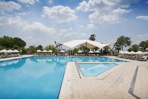 Uma grande piscina ao ar livre com espreguiçadeiras e guarda-sóis, cercada por árvores e um céu claro.