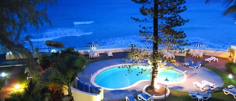 Área de piscina com espreguiçadeiras e guarda-sóis, cercada por vegetação e com vista para o oceano.