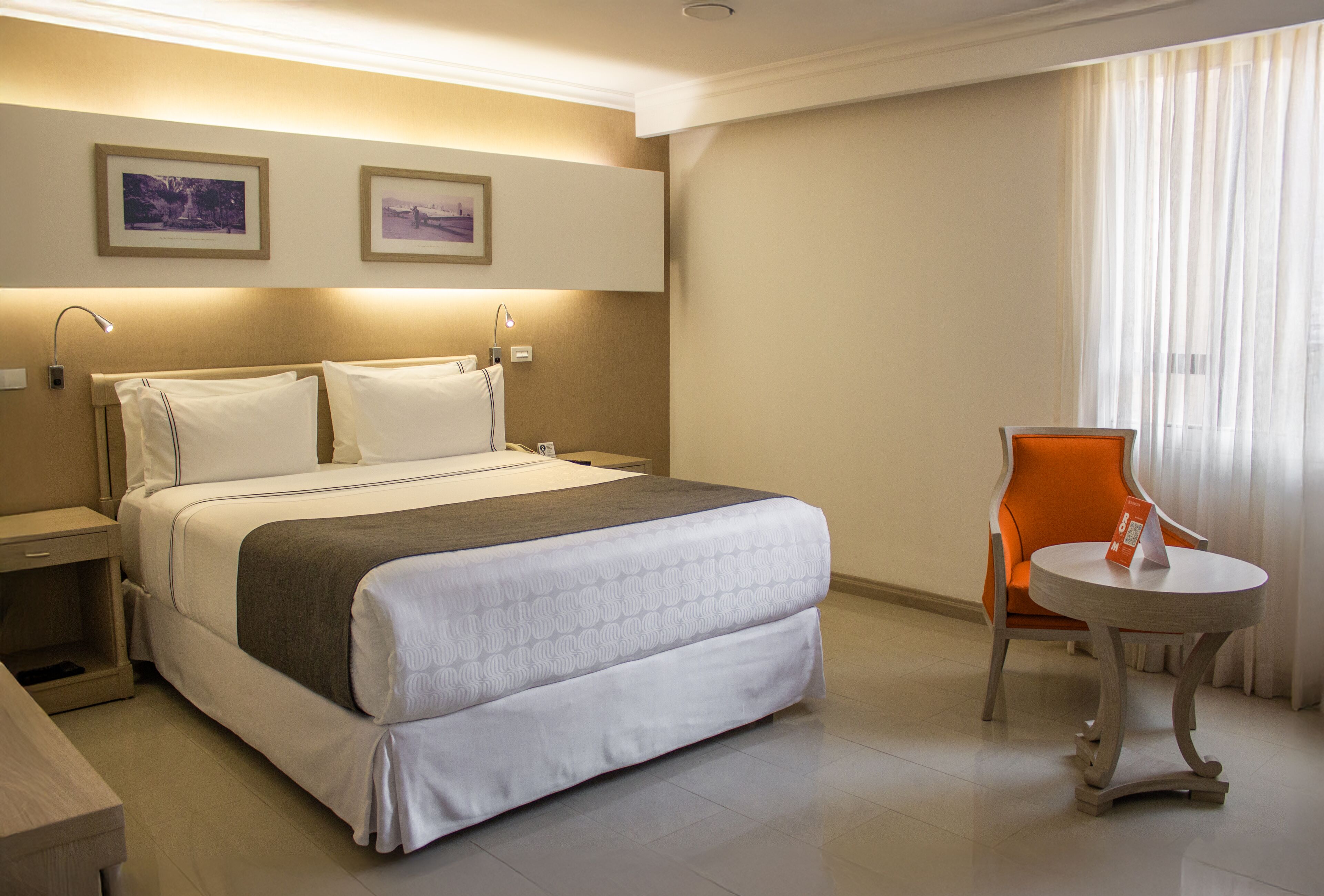 Quarto de hotel com uma cama, mesas de cabeceira, uma cadeira, uma mesinha e duas fotografias emolduradas na parede.