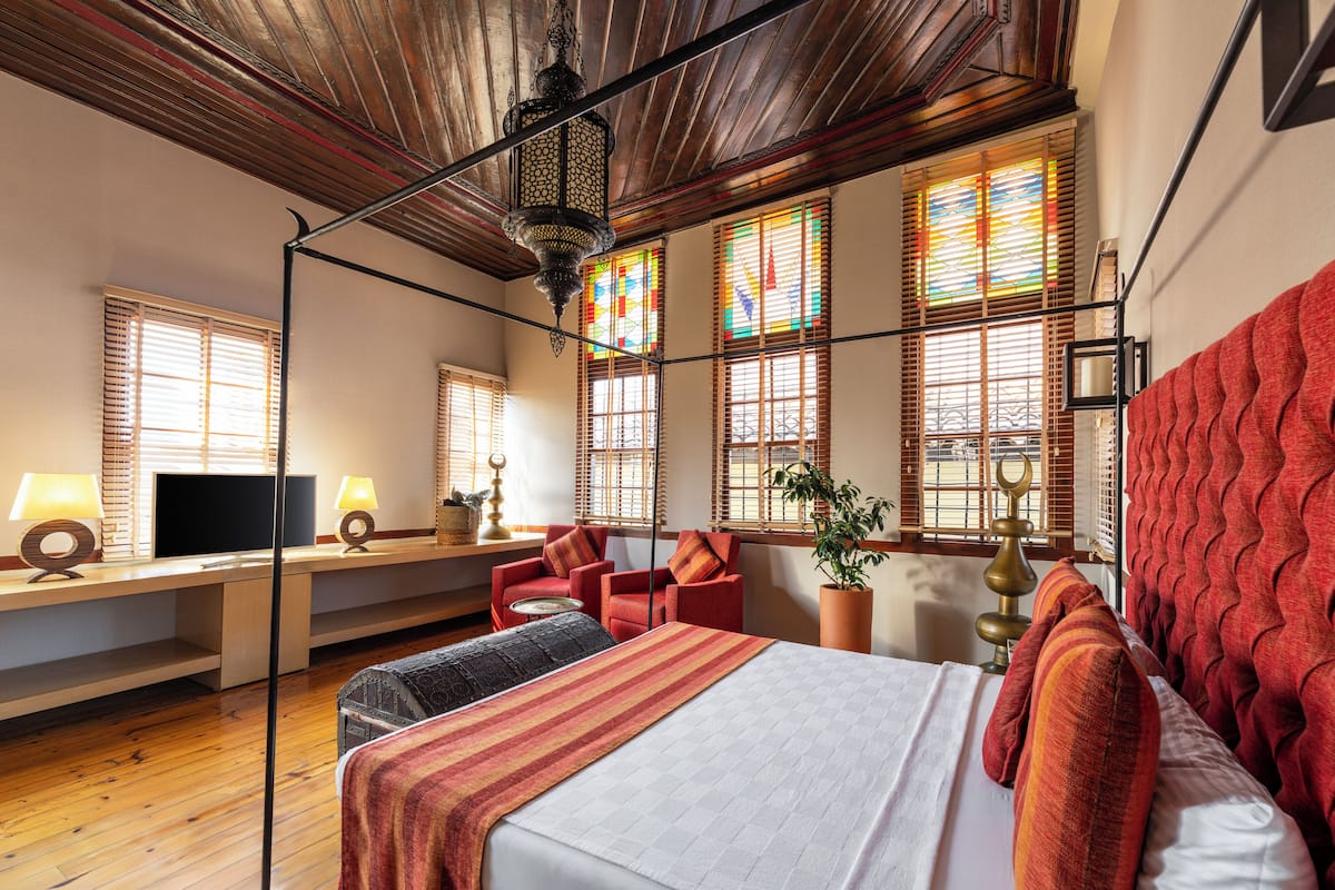 Quarto de hotel com teto de madeira, cama com cabeceira vermelha, escrivaninha com televisão e janela com vitrais.