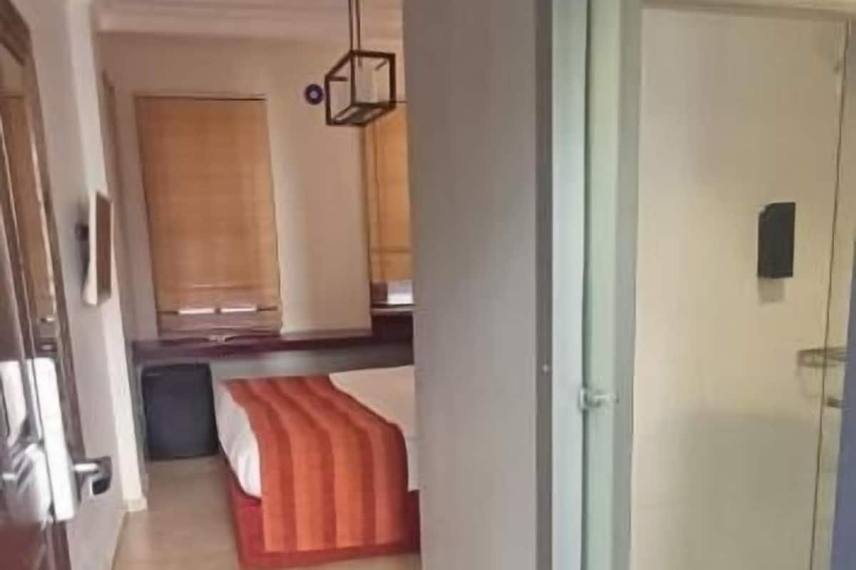 Corredor que leva a um quarto com porta de vidro para o chuveiro, cama com cabeceira laranja e área de cozinha ao fundo.