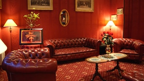 Um quarto com carpete vermelho estampado, um sofá de couro marrom, uma mesa redonda pequena com um vaso de flores e quadros nas paredes.