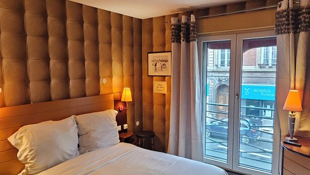 Quarto de hotel com uma cama grande, duas luminárias de cabeceira, um quadro na parede e vista da rua através de uma porta de vidro.