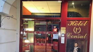 Entrada de hotel com fachada em vermelho e dourado, porta de vidro e retrato emoldurado de uma figura histórica.