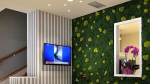 Um lobby moderno com um sofá de couro marrom, uma cadeira branca e uma pequena mesa redonda. Uma televisão de tela plana está fixada na parede, e um espelho reflete um vaso com flores.