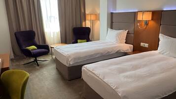 Quarto de hotel com duas camas, uma escrivaninha, uma cadeira e uma luminária.