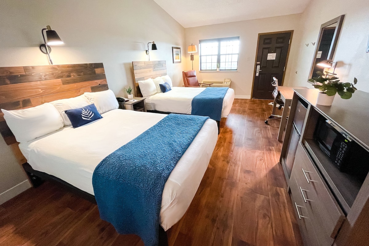Quarto de hotel com duas camas, piso de madeira, uma escrivaninha com cadeira, micro-ondas e uma planta em vaso.
