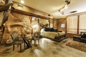 Quarto com uma cama grande, uma mesa de vidro, duas cadeiras estampadas e um detalhe decorativo na parede.