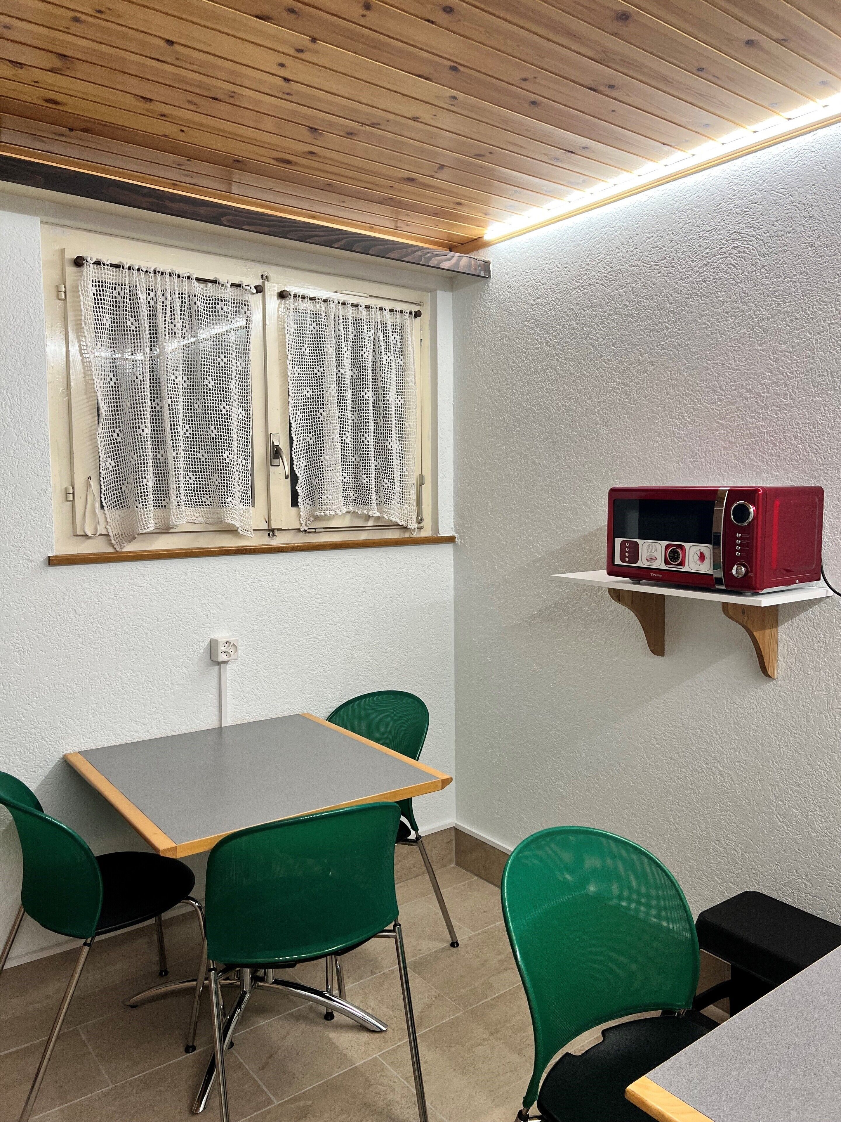 Um quarto com uma mesa e cadeiras, uma janela com cortina de crochê, um rádio vermelho numa prateleira e teto de madeira.