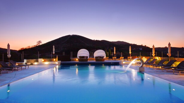 Piscina com espreguiçadeiras e guarda-sóis, cercada por um muro de pedra e vista para uma colina ao entardecer.