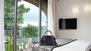 Quarto com uma janela ampla, uma cama, uma TV e vista para o verde.