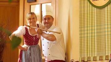 Um homem e uma mulher com vestes tradicionais, possivelmente em uma cozinha ou sala de jantar, com uma fileira de garrafas de vinho ao fundo.
