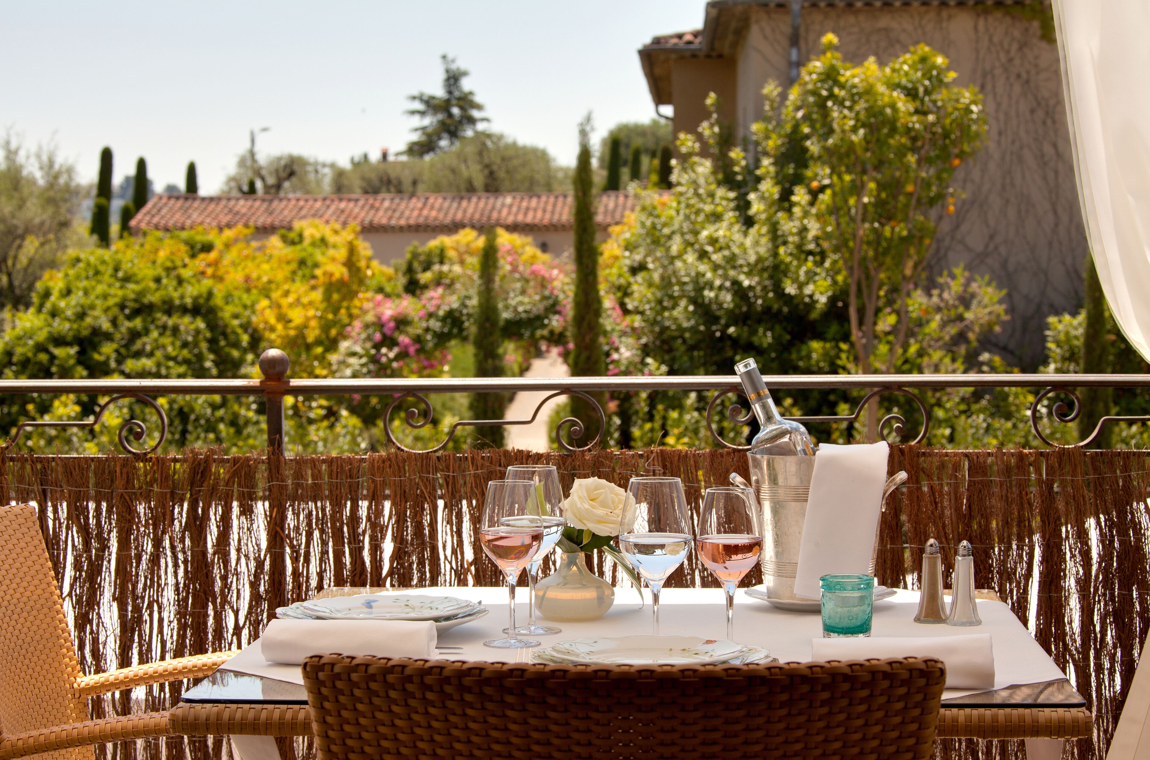 Mesa posta para dois, com taças de vinho, um vaso com uma única rosa branca e um jarro numa varanda com vista para um jardim com diversas árvores e um edifício com telhado de cerâmica.
