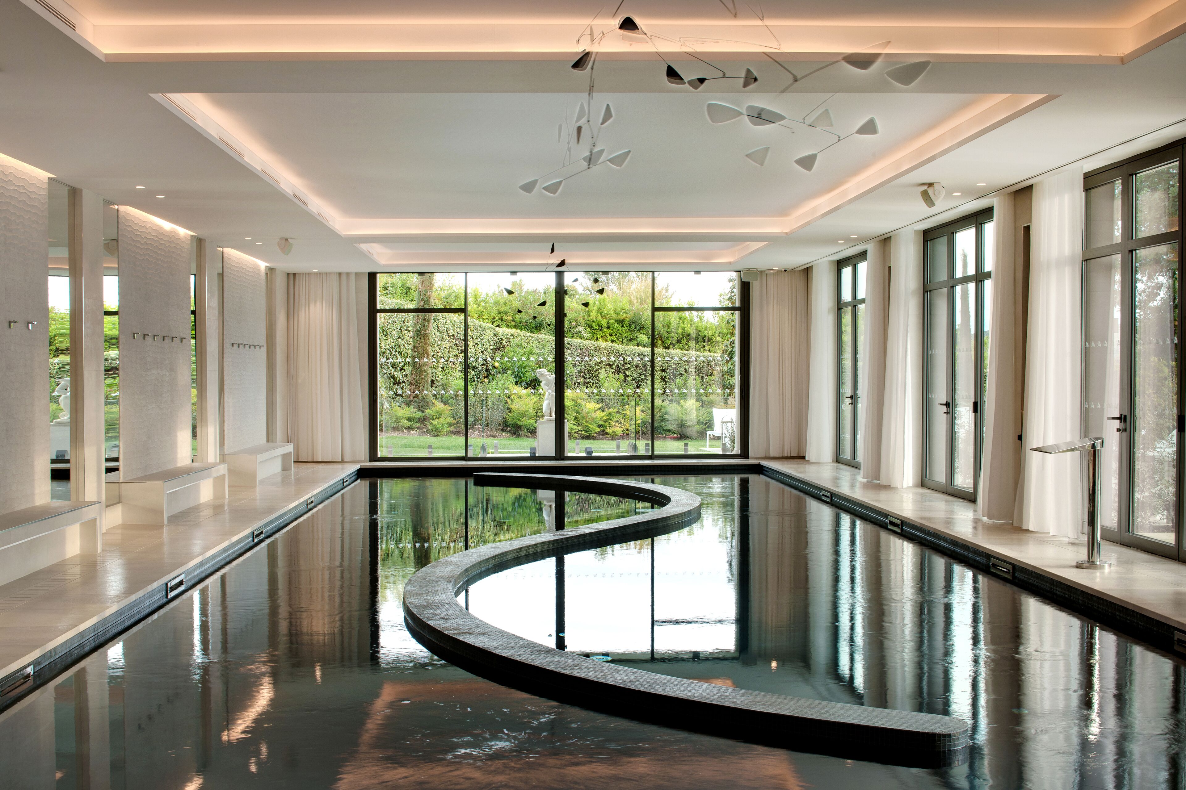 Piscina interior moderna com design curvo, grandes janelas e vista para o verde lá fora.