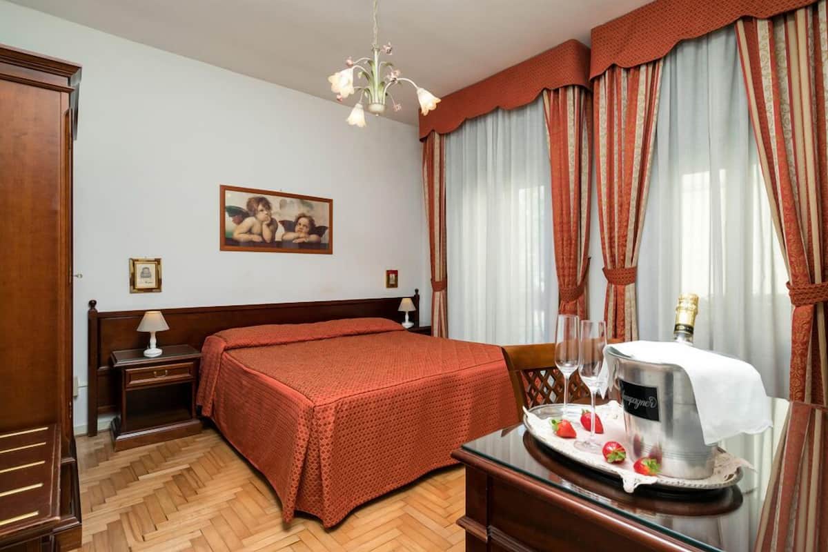Quarto de hotel com cama de madeira, mesa de cabeceira, escrivaninha com uma garrafa de champanhe e um quadro na parede.