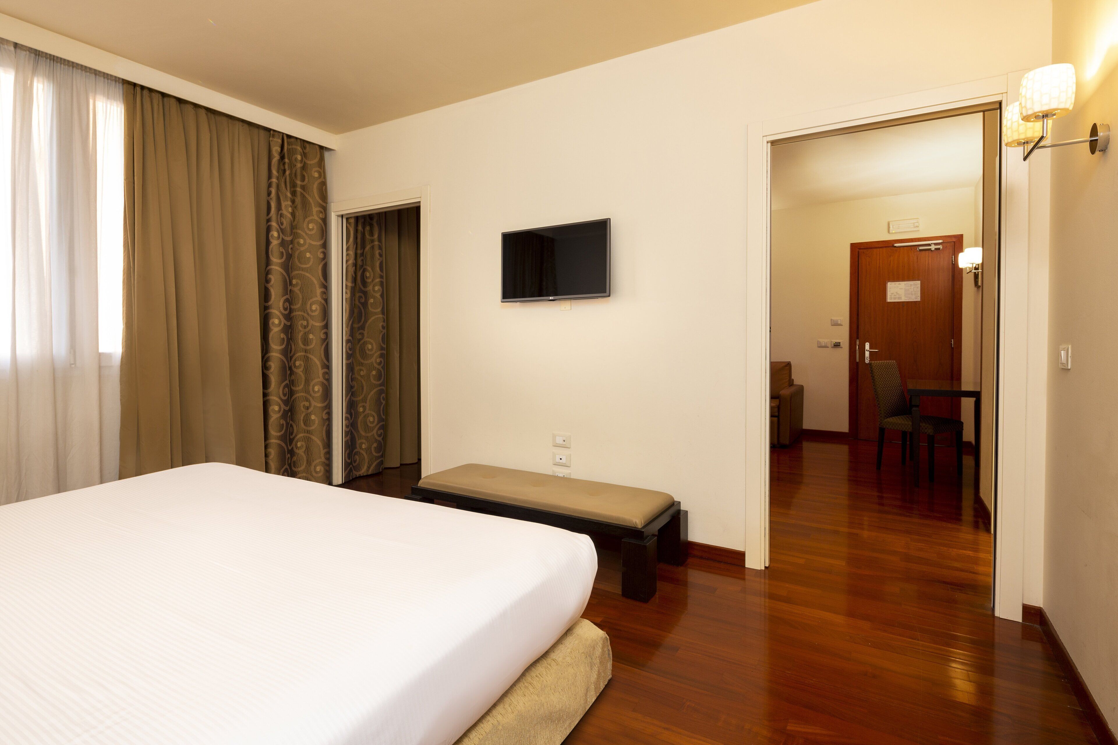 Quarto de hotel com uma cama, um banco, uma televisão e uma porta que dá para outro espaço.