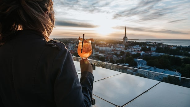 Uma pessoa segurando um copo com canudo, apreciando a vista de uma paisagem urbana com uma torre de igreja imponente ao pôr do sol.